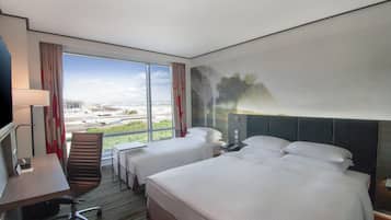 Quarto de hotel com duas camas, uma mesa, uma cadeira e uma grande janela com vista para a cidade.