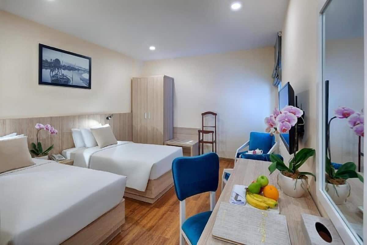 Quarto de hotel com duas camas, uma escrivaninha com frutas, uma cadeira, uma televisão e uma janela com uma planta orquídea.