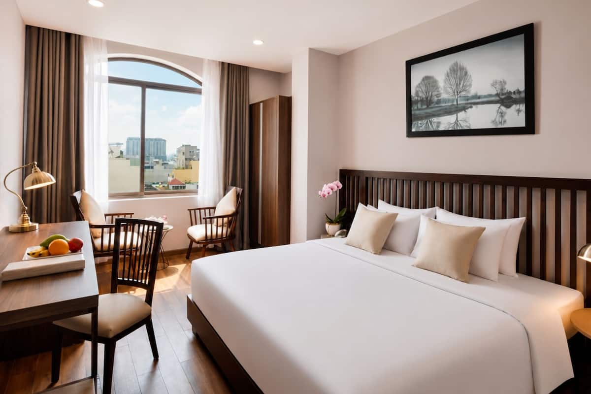 Quarto de hotel com uma cama grande, uma escrivaninha com luminária, uma cadeira, uma janela com cortinas e um quadro na parede.