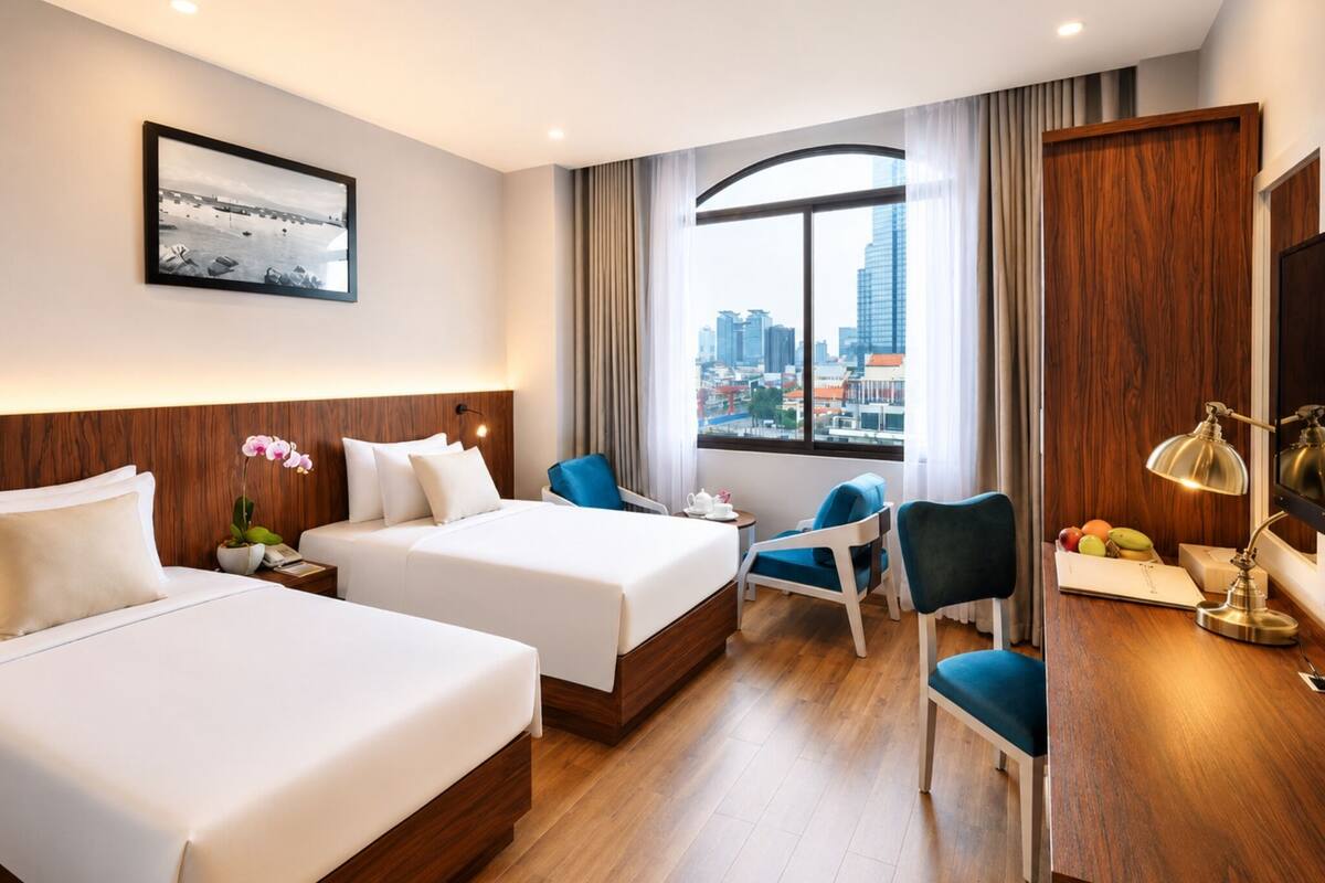 Quarto de hotel com duas camas, uma escrivaninha com luminária, uma cadeira, vista para a cidade e uma imagem emoldurada na parede.