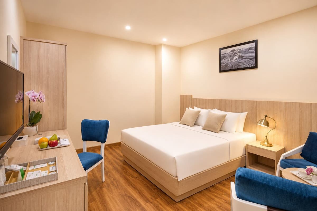 Quarto de hotel com uma cama, uma escrivaninha com cadeira, uma mesinha de apoio com abajur e um quadro na parede.