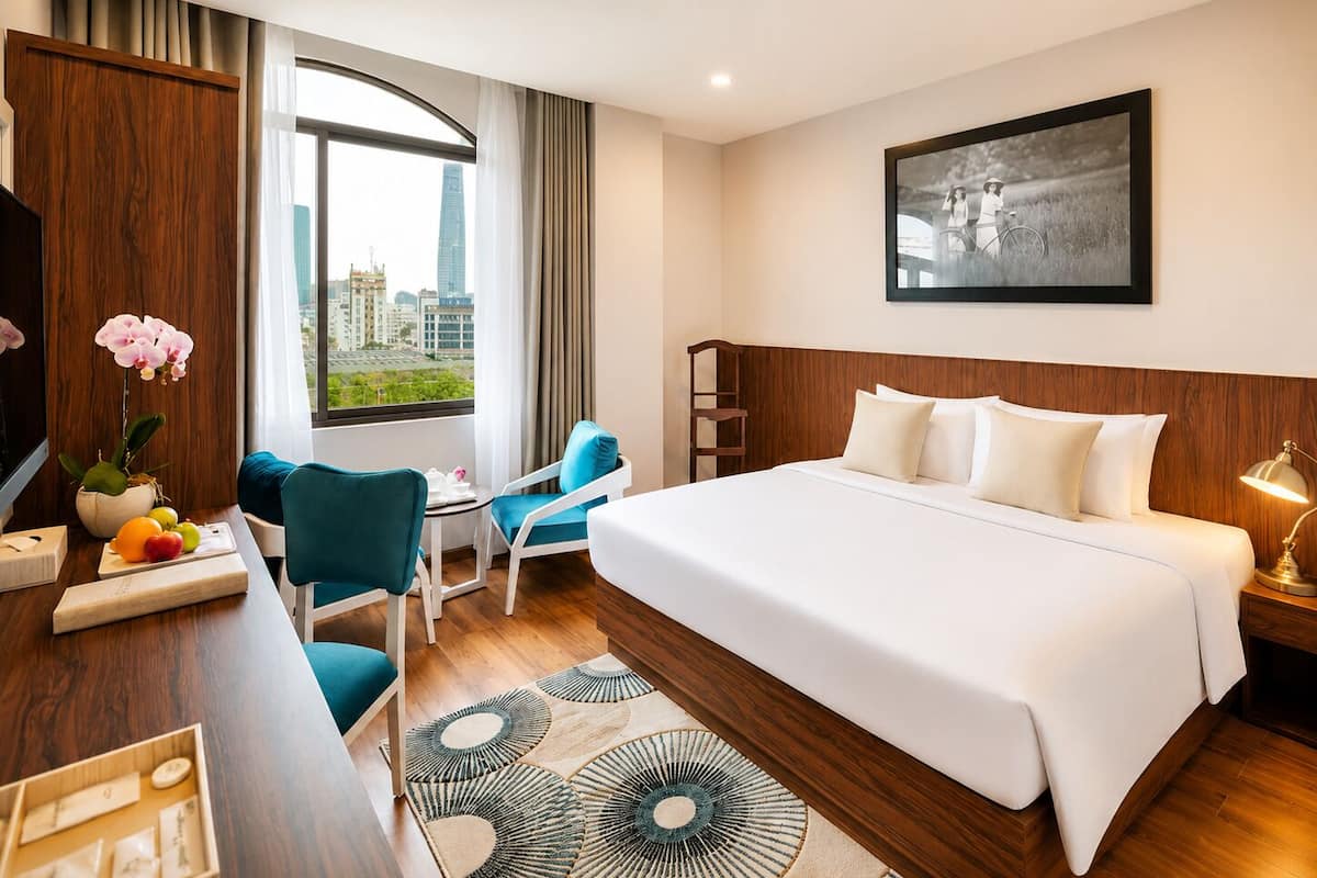 Quarto de hotel com uma cama grande, uma escrivaninha com cadeira, uma mesinha, uma poltrona, uma janela com vista para edifícios e um quadro na parede.