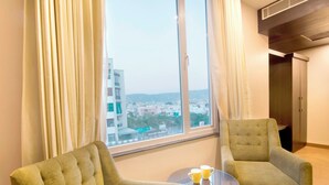 Quarto com duas poltronas verdes, uma mesinha de vidro com duas chávenas amarelas e uma janela ampla com vista para a cidade.