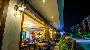 Uma área de refeições ao ar livre com mesas e cadeiras de madeira, um letreiro de neon e vista para a rua à noite.