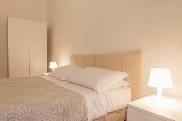 Cama bem arrumada com colcha estampada, abajur e guarda-roupa em um quarto simples e arejado.