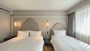 Quarto de hotel com duas camas, uma mesinha redonda e duas luminárias fixadas na parede.