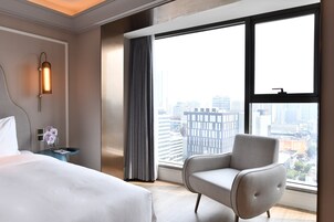 Quarto de hotel com cama, cadeira, mesa de cabeceira, luminária, janela com vista para a cidade e cortina.