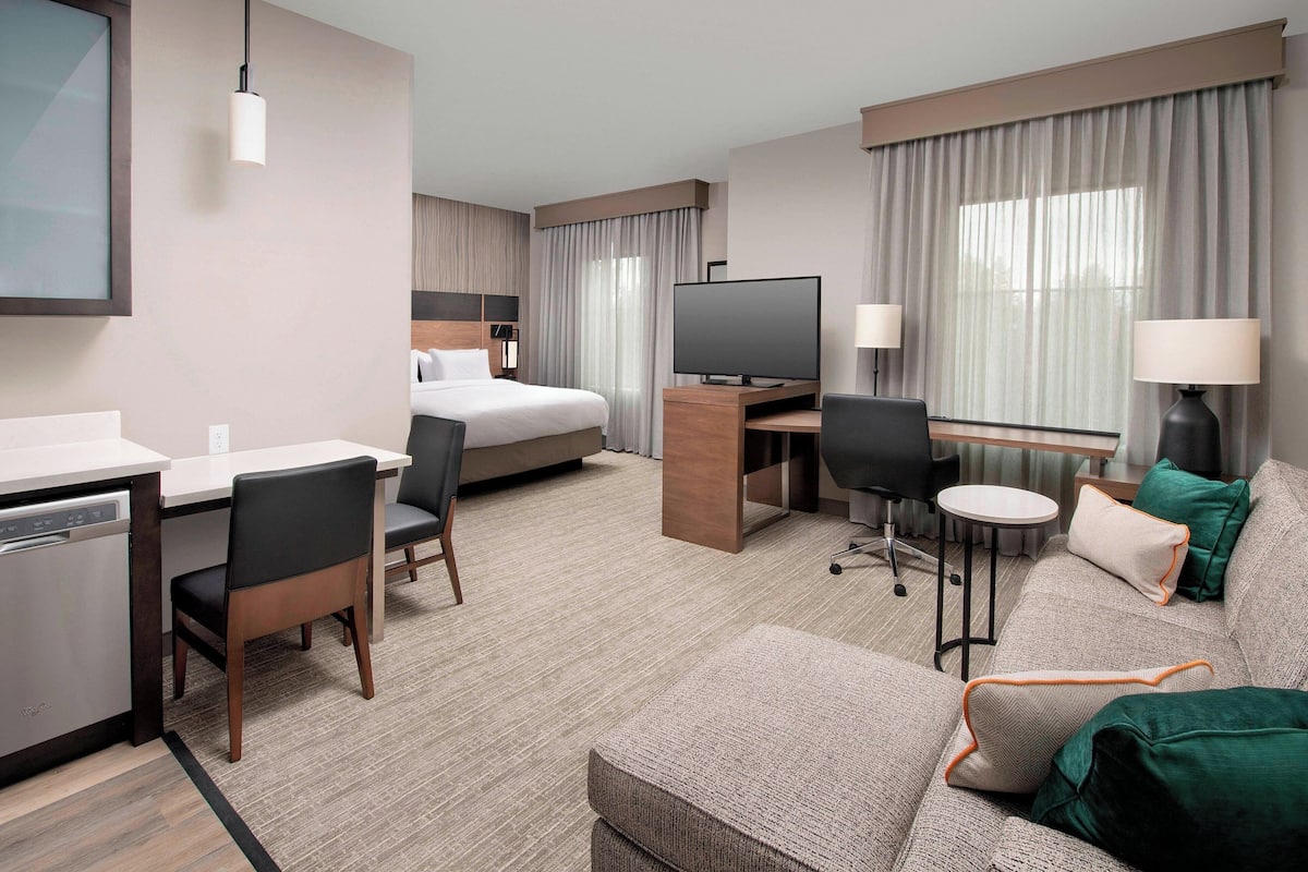 Quarto de hotel moderno com cama, escrivaninha com televisão, área de jantar com cadeira e mesa, e sofá com almofadas.