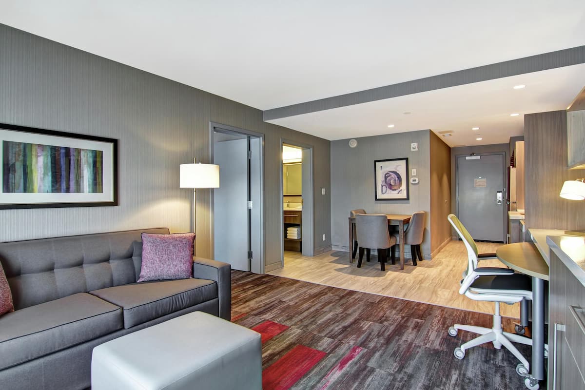 Quarto de hotel moderno com sofá cinzento, uma mesa pequena, escrivaninha com cadeira e área de refeições com cadeiras.