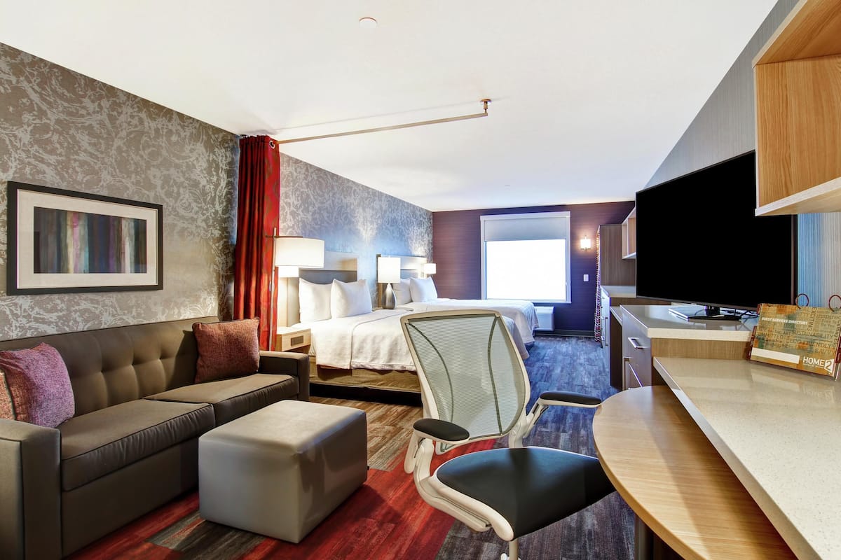 Quarto de hotel com sofá, banquinho, poltrona, cama, escrivaninha, televisão e uma grande janela.