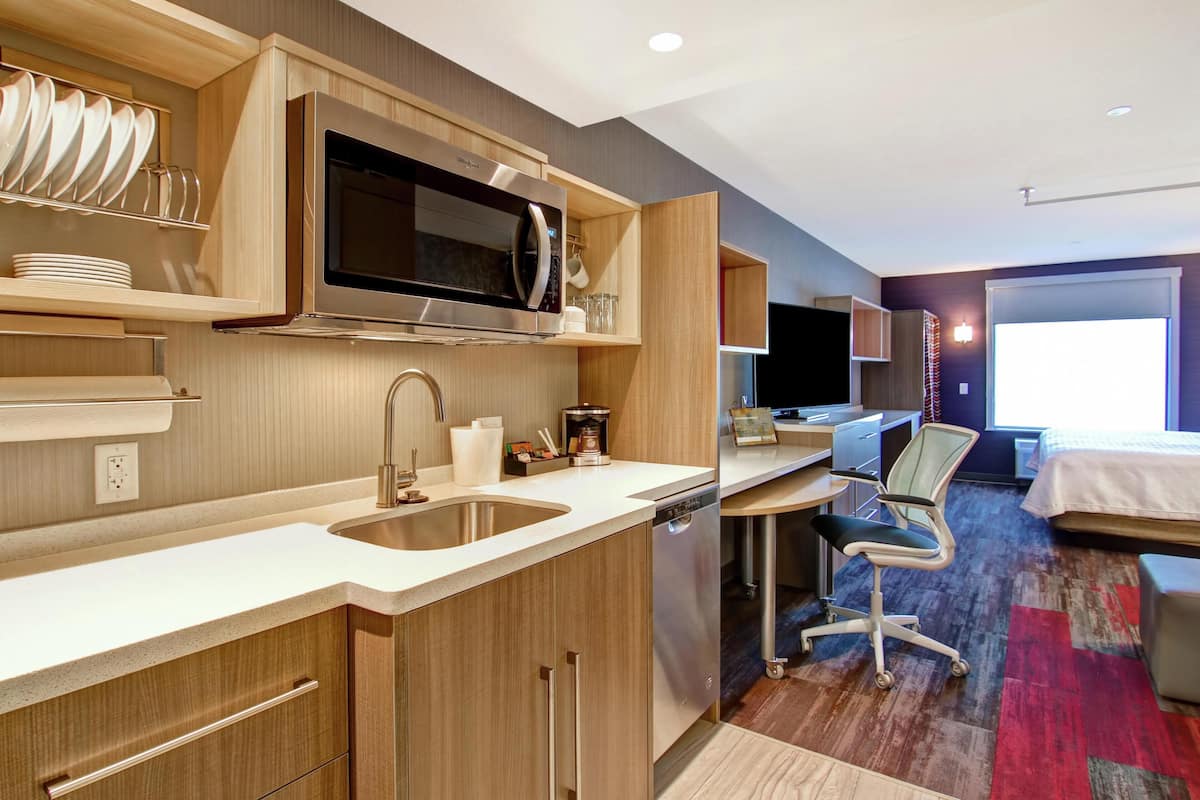 Quarto de hotel moderno com cozinha compacta, uma janela grande, cama e escrivaninha.