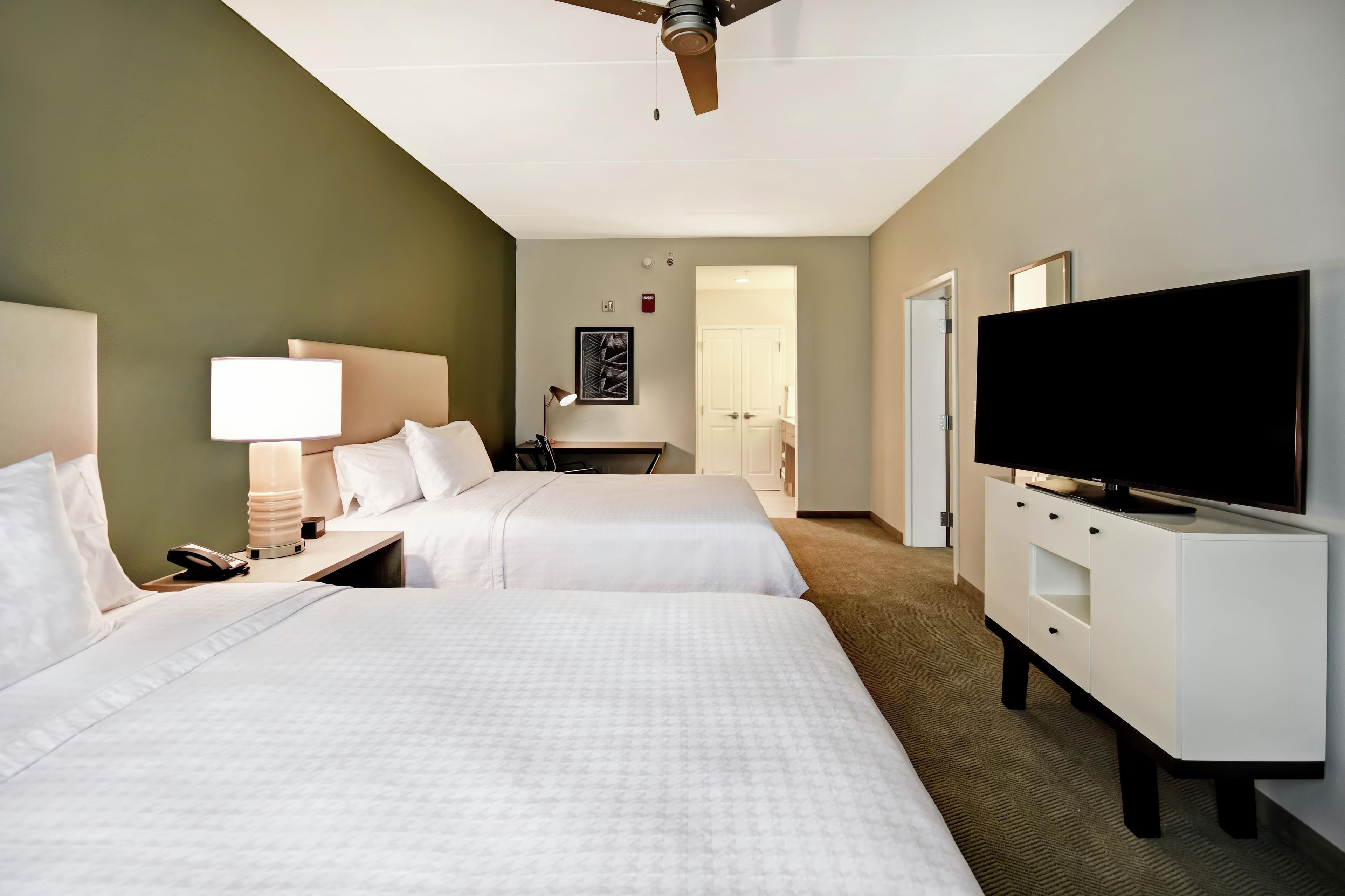 Quarto de hotel com duas camas, televisão, escrivaninha com abajur e ventoinha no teto.
