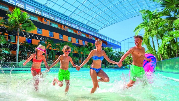 Área de piscina de hotel com água cristalina, palmeiras e um edifício com cobertura de vidro identificado como 'HOTEL VICTORY'.