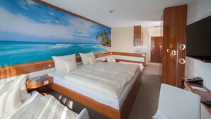 Quarto de hotel com uma cama grande, uma escrivaninha, uma cadeira, uma TV e um quadro com uma paisagem de praia.