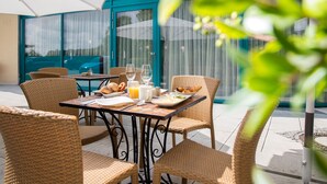 Uma área de refeições com cadeiras e mesas de vime, um buffet com comida e bebidas, e um grande guarda-sol oferecendo sombra.