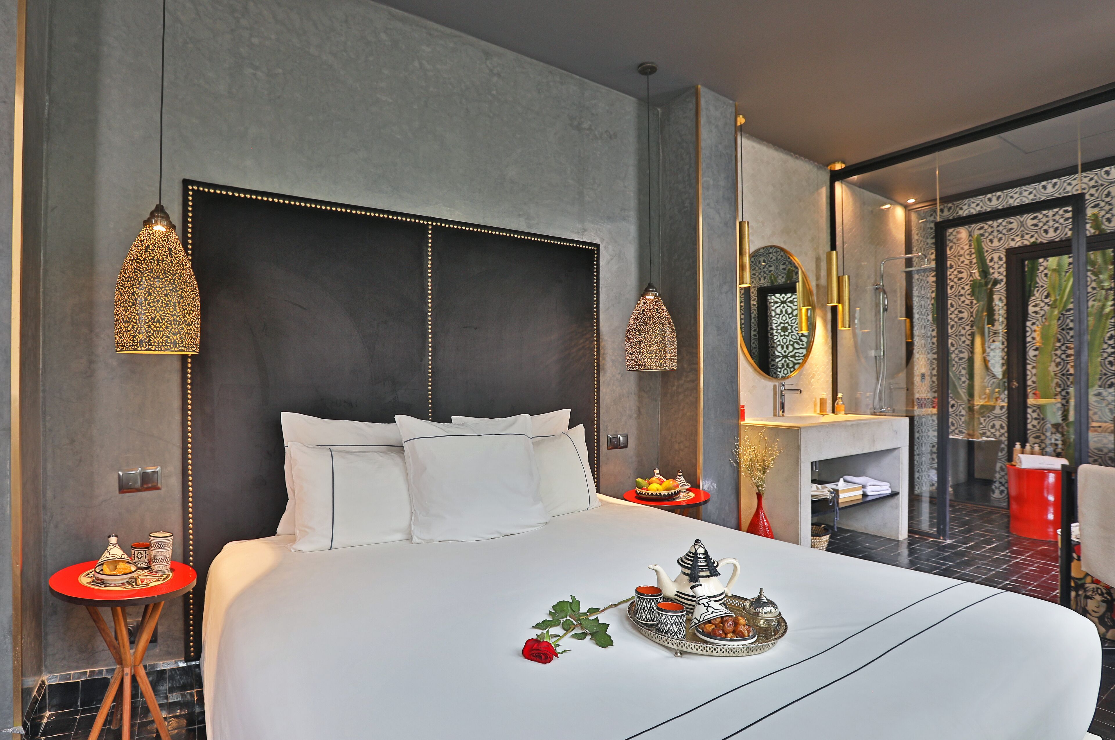 Quarto de hotel moderno com uma cama grande, mesa de cabeceira com bandeja de comida e banheiro com espelho grande e azulejos estampados.