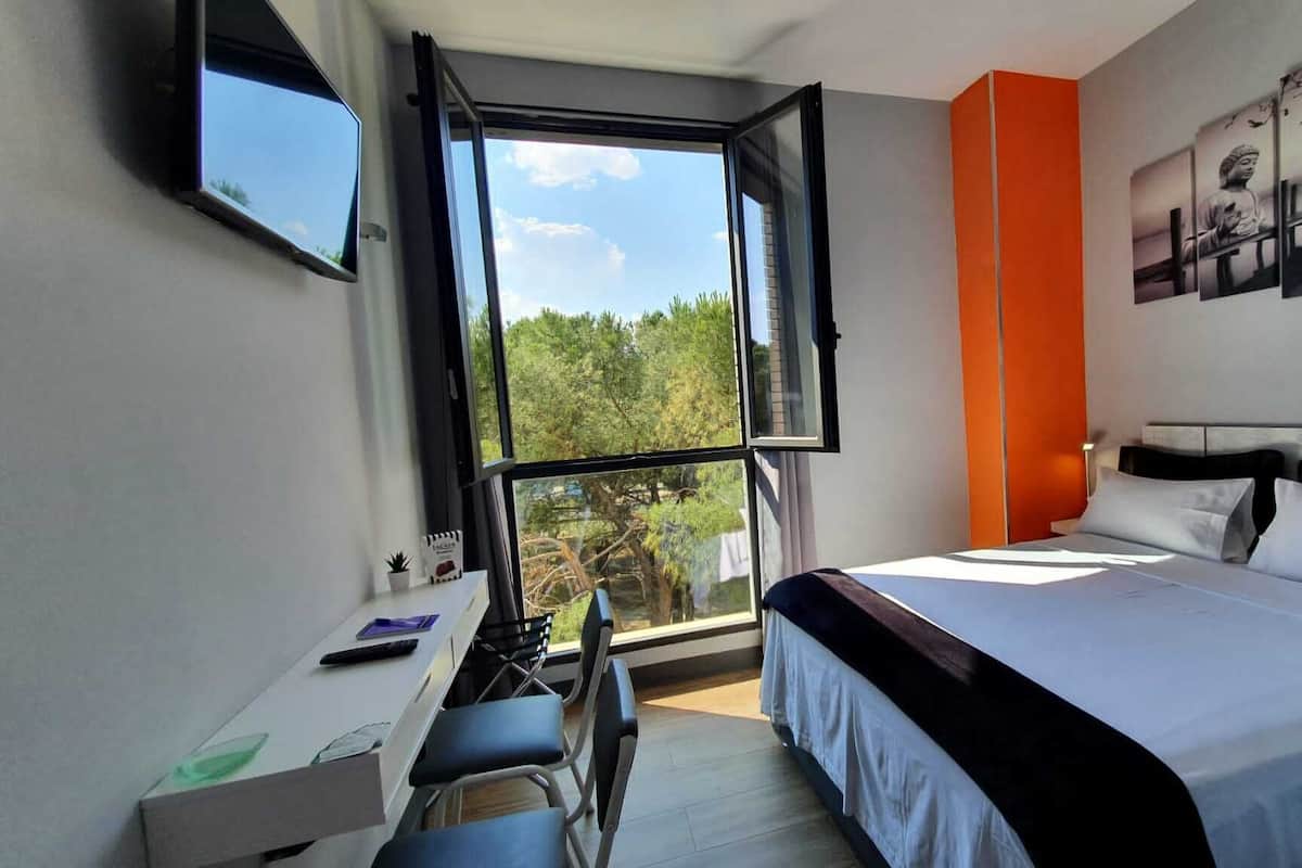 Quarto de hotel moderno com uma cama grande, uma escrivaninha e uma cadeira. Há uma janela com vista para árvores e um céu limpo.