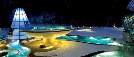 Área externa coberta por neve, com piscina aquecida central, estrutura de vidro e montanhas ao fundo.