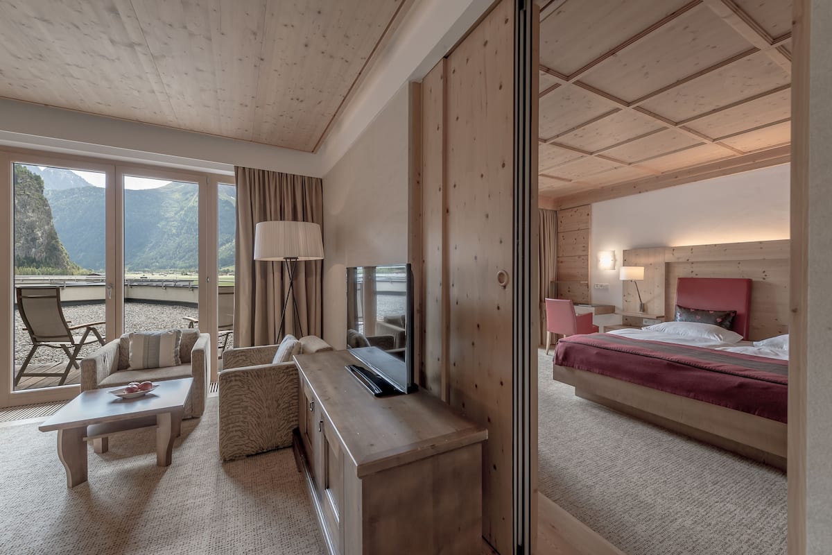 Quarto de hotel com uma grande porta de vidro deslizante, cama, escrivaninha com cadeira, sofá e televisão.