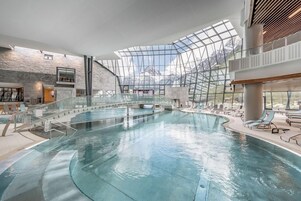 Piscina interna com teto de vidro, espreguiçadeiras e vista para montanhas.