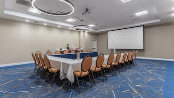 Sala de conferência com uma longa mesa, cadeiras, tela de projetor e piso coberto por carpete.