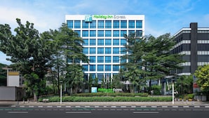 Um hotel Holiday Inn Express com design moderno, apresentando um logotipo destacado, grandes janelas e uma área frontal ajardinada com árvores e um banco.