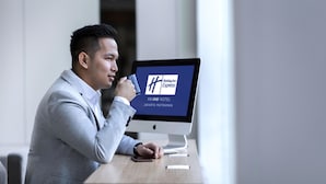 Um homem de terno sentado em uma mesa em frente a um computador que exibe uma interface de reserva de hotel.