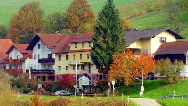 Uma fileira de casas tradicionais em estilo europeu, com telhados inclinados, um edifício central com varanda, e um morro verde ao fundo com árvores em tons de outono.