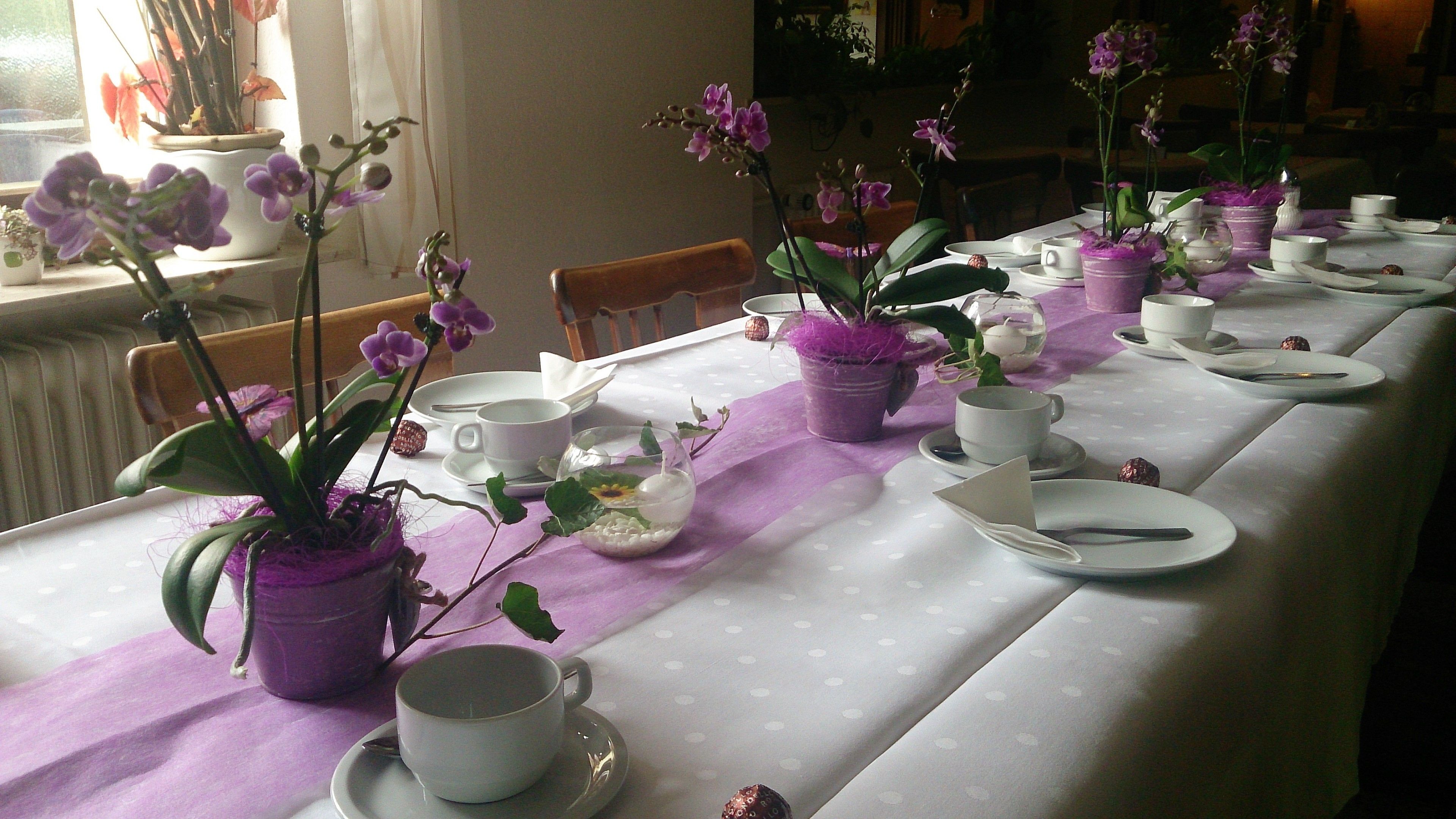 Uma mesa de jantar posta com pratos brancos, chávenas e talheres, decorada com orquídeas roxas em vasos.
