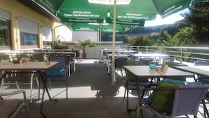 Um pátio externo com mesas e cadeiras, guarda-sóis verdes e vista para a paisagem ao redor.