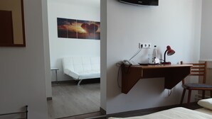 Quarto de hotel com cama, escrivaninha com luminária, cadeira, televisão fixada na parede e um quadro sobre a escrivaninha.