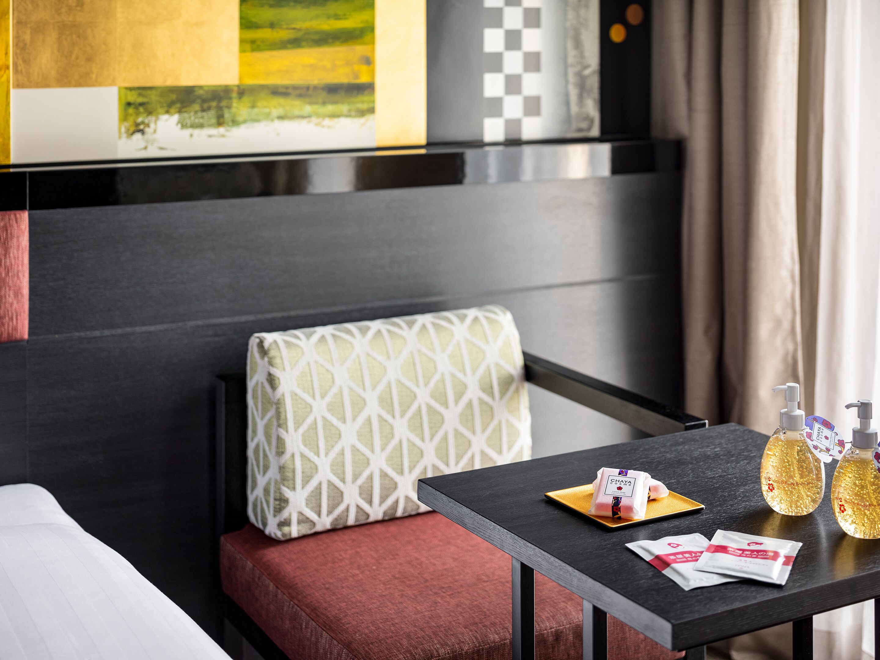 Quarto de hotel com cama, sofá, pequena mesa com artigos de higiene e almofada com padrão geométrico.