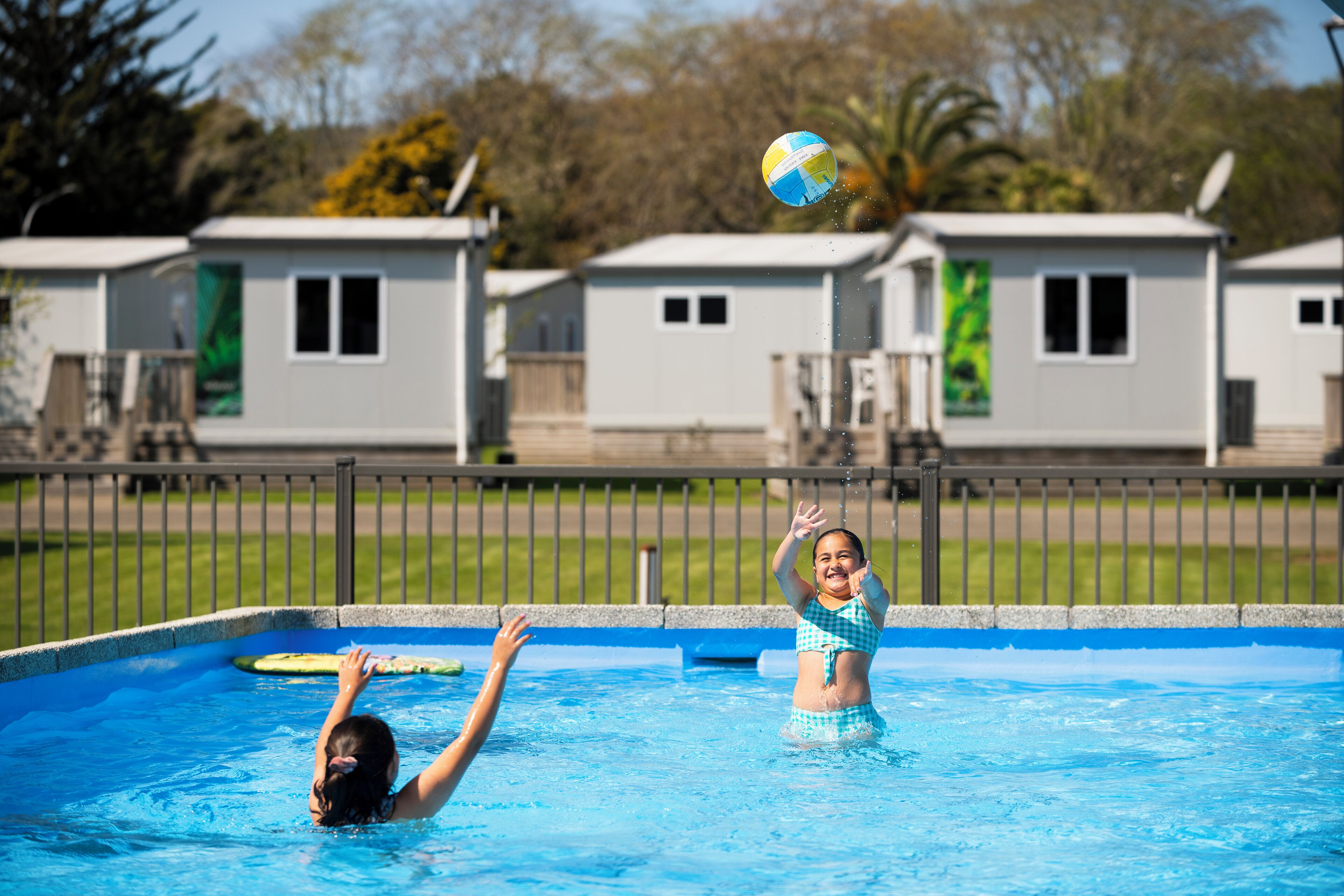 Piscina com duas pessoas brincando com uma bola. Edifícios pequenos ao fundo.