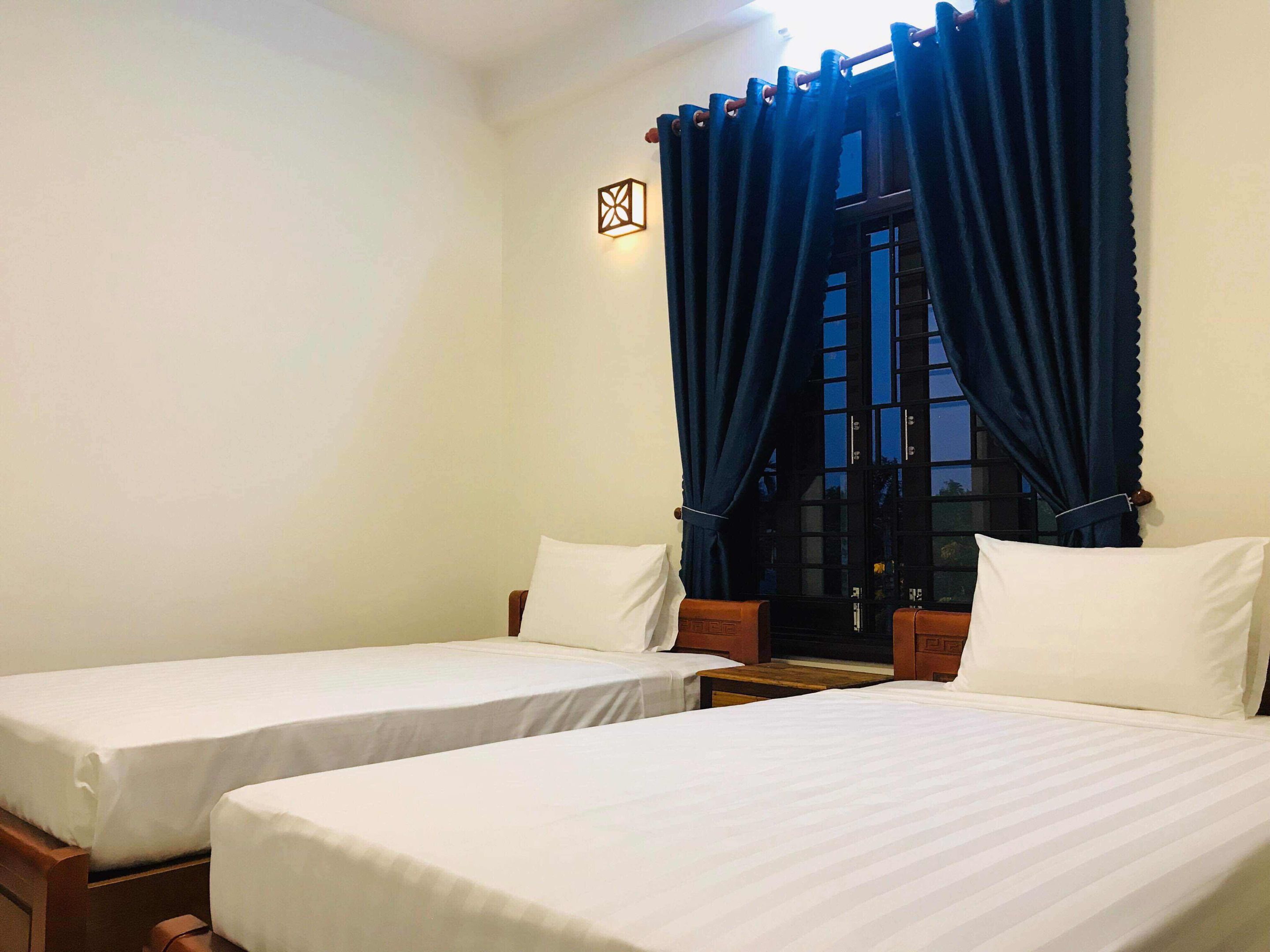Quarto de hotel com duas camas de solteiro, uma pequena mesa de cabeceira de madeira, uma janela com cortinas escuras e uma luminária fixada na parede.
