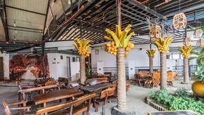 Um restaurante com bancos e mesas de madeira, palmeiras decorativas e um mural na parede.