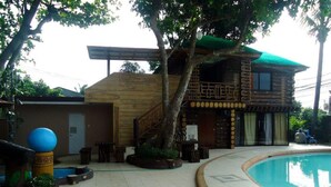 Um edifício de madeira com piscina, rodeado por árvores e um pátio com azulejos.