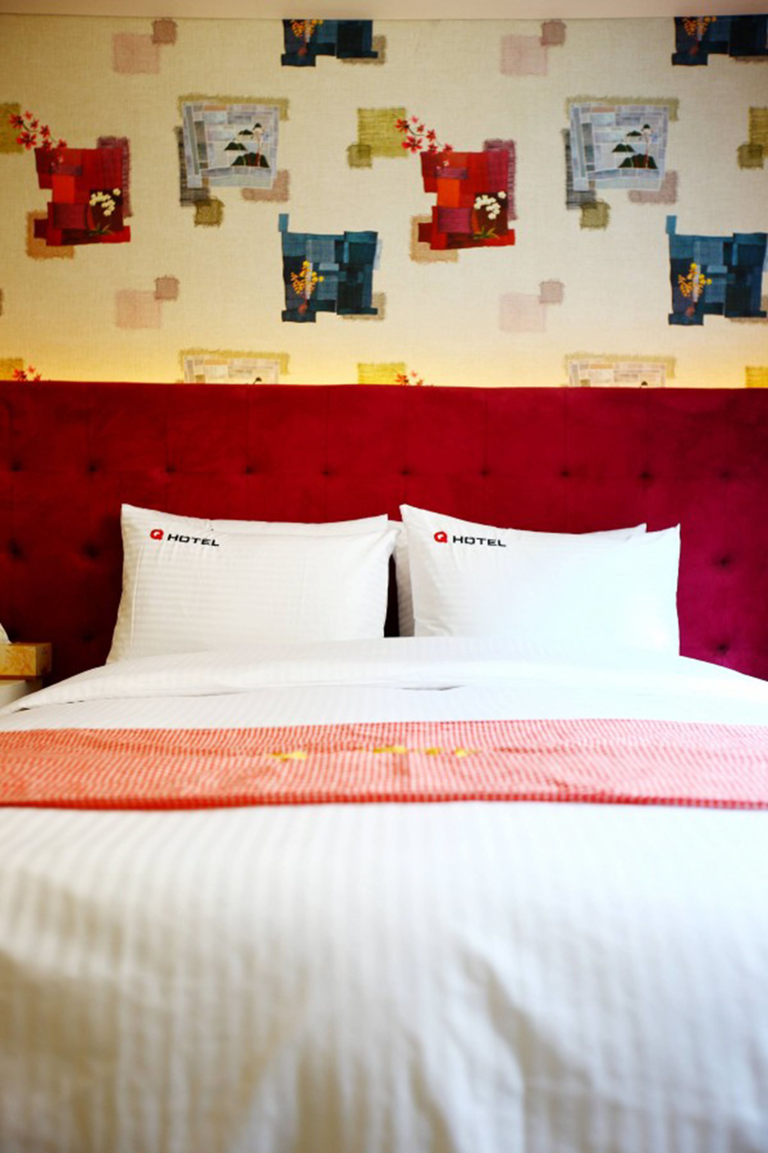 Quarto de hotel com cabeceira estofada vermelha, almofadas brancas com o logotipo do hotel e uma parede estampada.