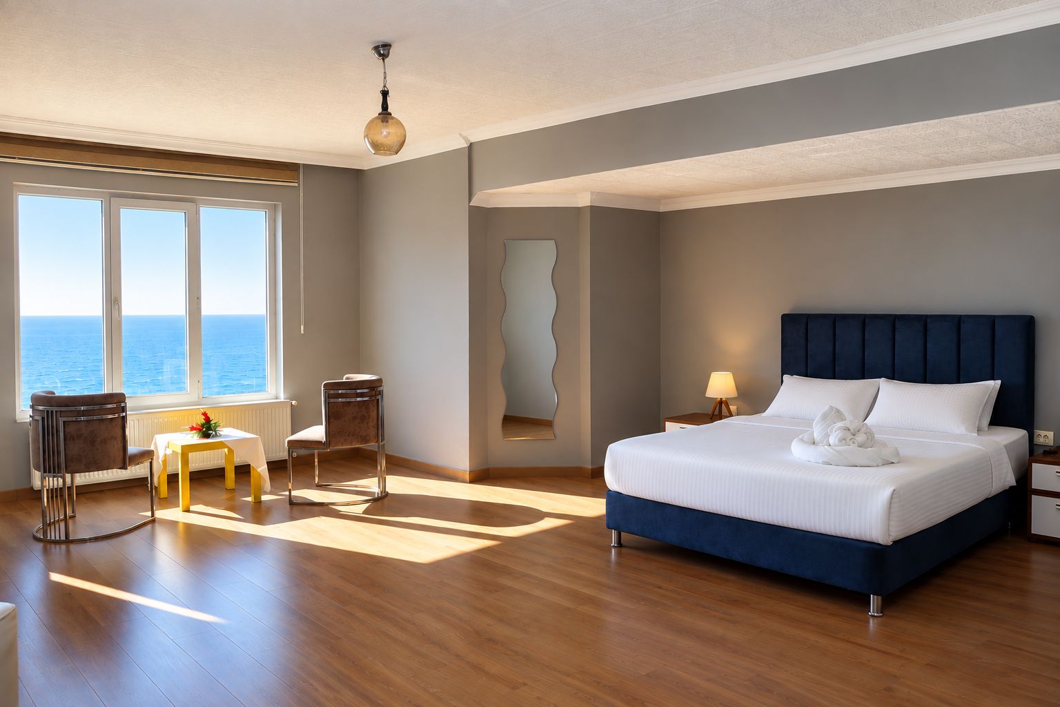 Quarto de hotel com uma cama grande, vista para o mar, área de refeições com mesa e cadeiras, e um espelho na parede.