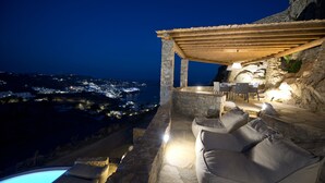 Um terraço de pedra com pérgola de madeira, área de estar ao ar livre e vista para uma cidade costeira à noite.