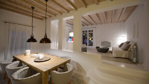 Uma sala de jantar com mesa de madeira, cadeiras e luminárias pendentes. Há um sofá e uma janela com cortinas ao fundo.