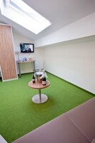 Quarto com carpete verde, mesa redonda com garrafas, TV na parede e painel de madeira.