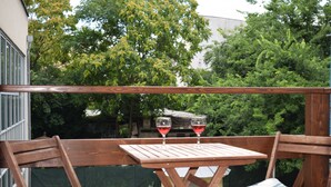 Um deck de madeira com uma mesa e duas cadeiras, dois copos de vinho tinto na mesa, e vista para árvores e um prédio ao fundo.