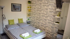 Quarto bem arrumado com cama, almofadas, abajur, quadro na parede e estante com livros e planta.
