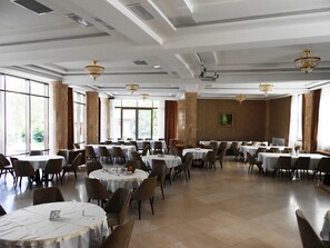 Uma ampla sala de banquetes com várias mesas redondas e cadeiras, janelas grandes e lustres elegantes.