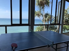 Mesa de pingue-pongue com rede em um quarto com janelas amplas dando para a água e palmeiras.