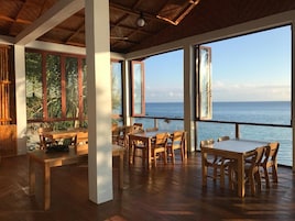 Uma sala de jantar ampla, com mesas e cadeiras de madeira, janelas grandes com vista para o mar e teto de madeira.