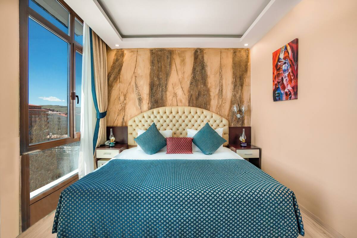 Quarto com uma cama grande, cabeceira e vista para a cidade através de uma janela ampla.