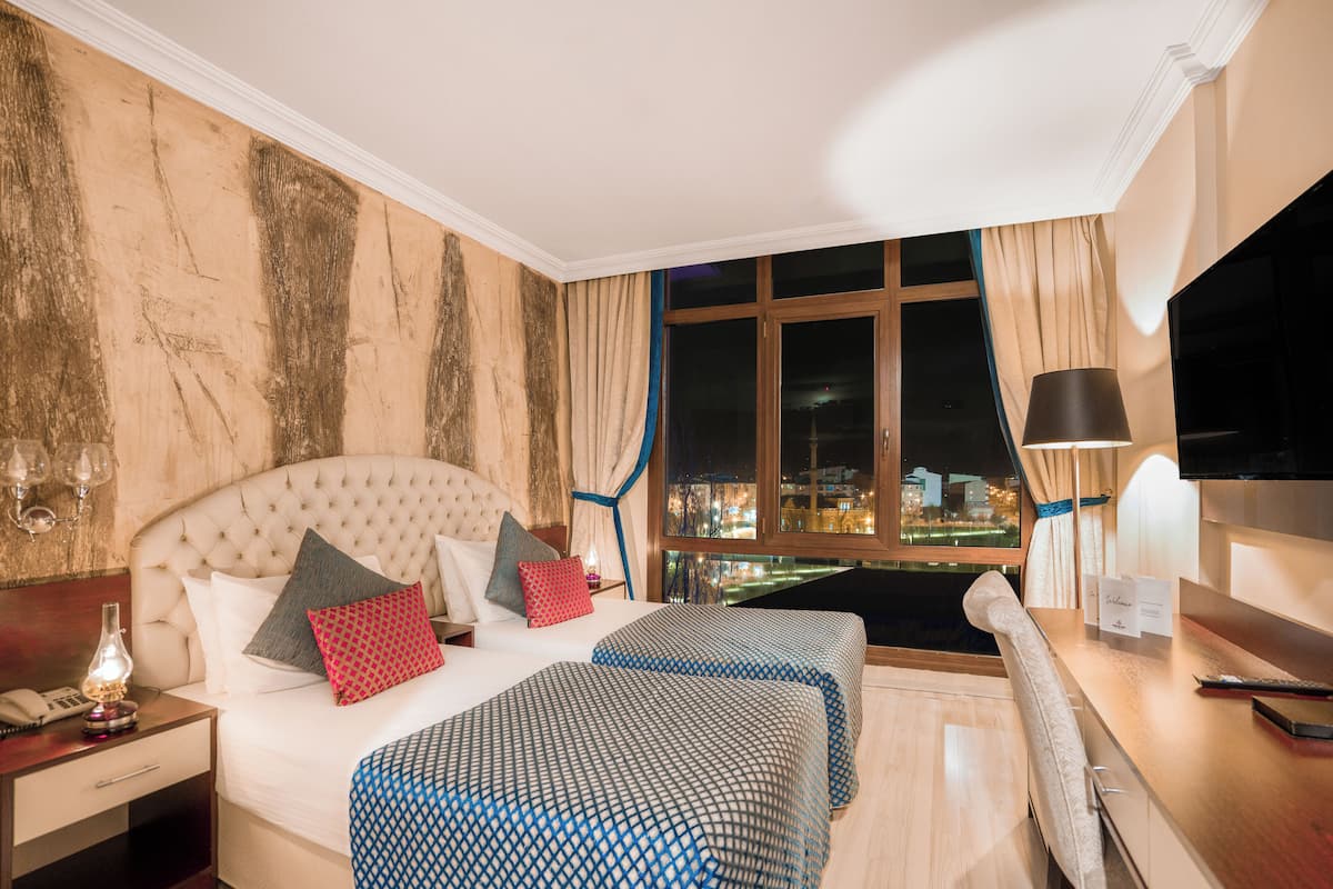 Quarto de hotel com uma cama grande, duas mesas de cabeceira, uma escrivaninha, uma cadeira e uma televisão.