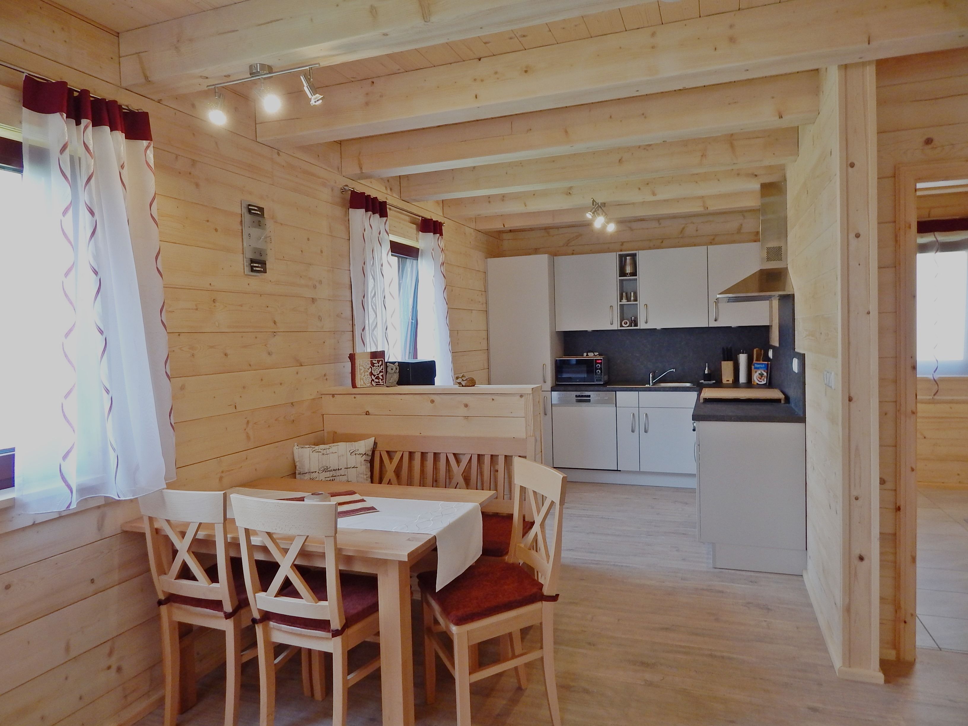 Interior de uma cabana de madeira com área de jantar, cozinha e janela com cortinas.