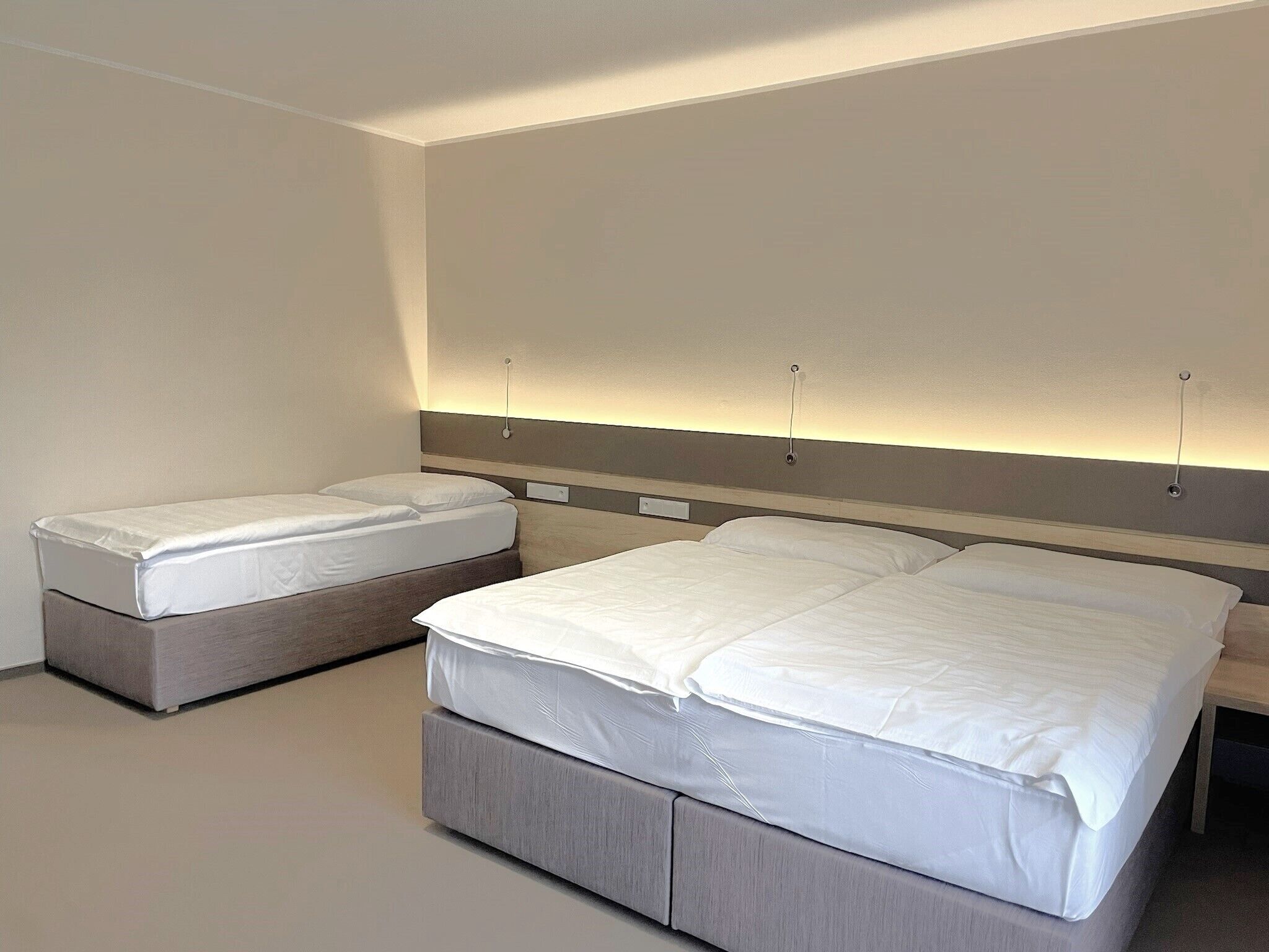 Quarto de hotel com duas camas, uma mesinha de cabeceira e uma luminária fixada na parede.
